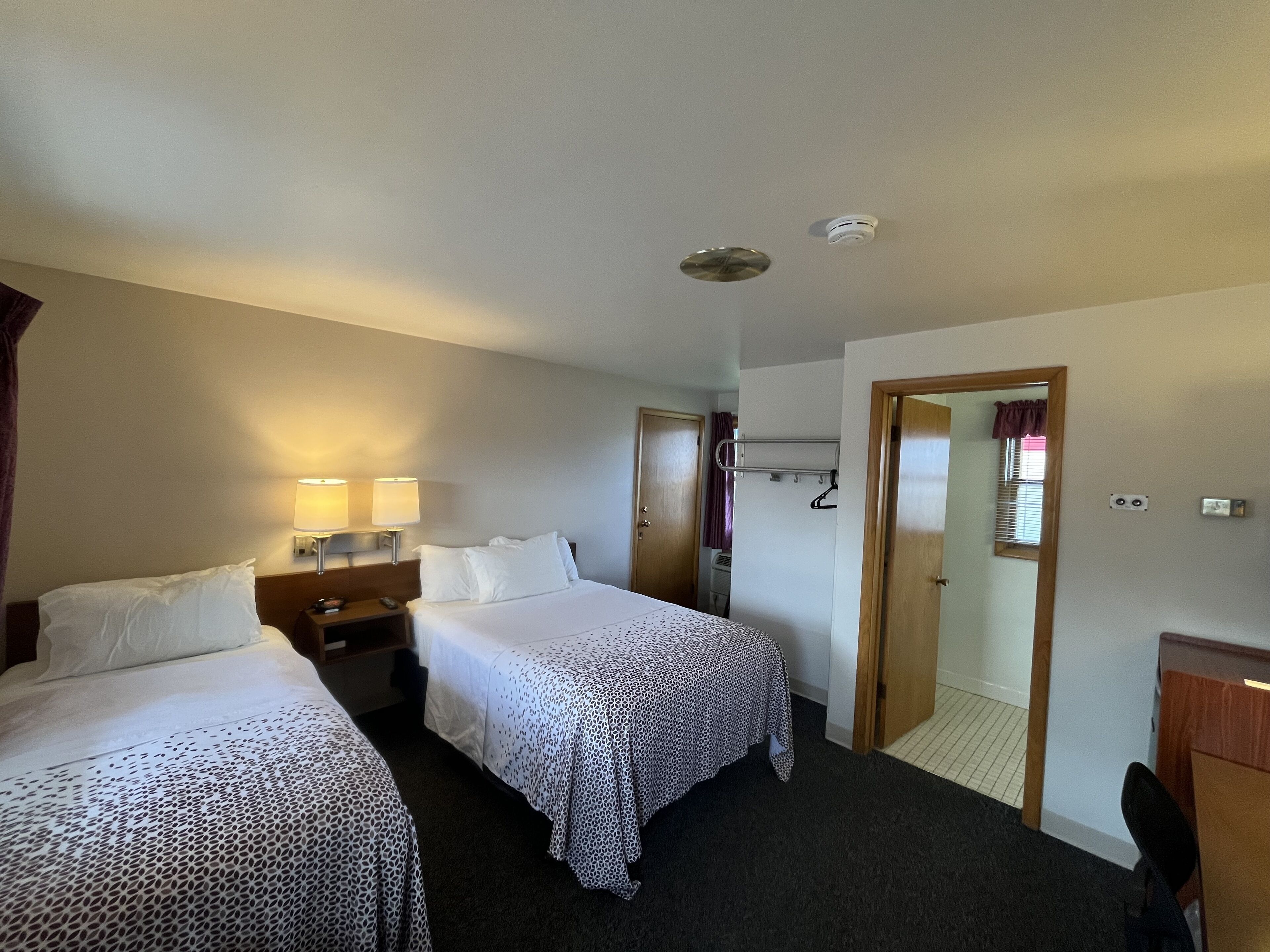 Quarto de hotel com duas camas, uma mesa de cabeceira, uma lâmpada, um espelho e uma porta que dá para outro cômodo.