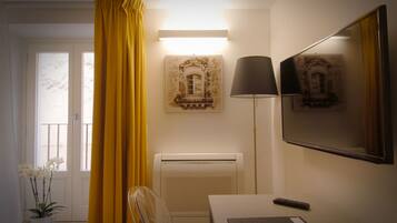 Quarto de hotel com uma cama, uma mesa com um laptop, uma luminária, uma TV, um radiador e uma janela com cortinas.