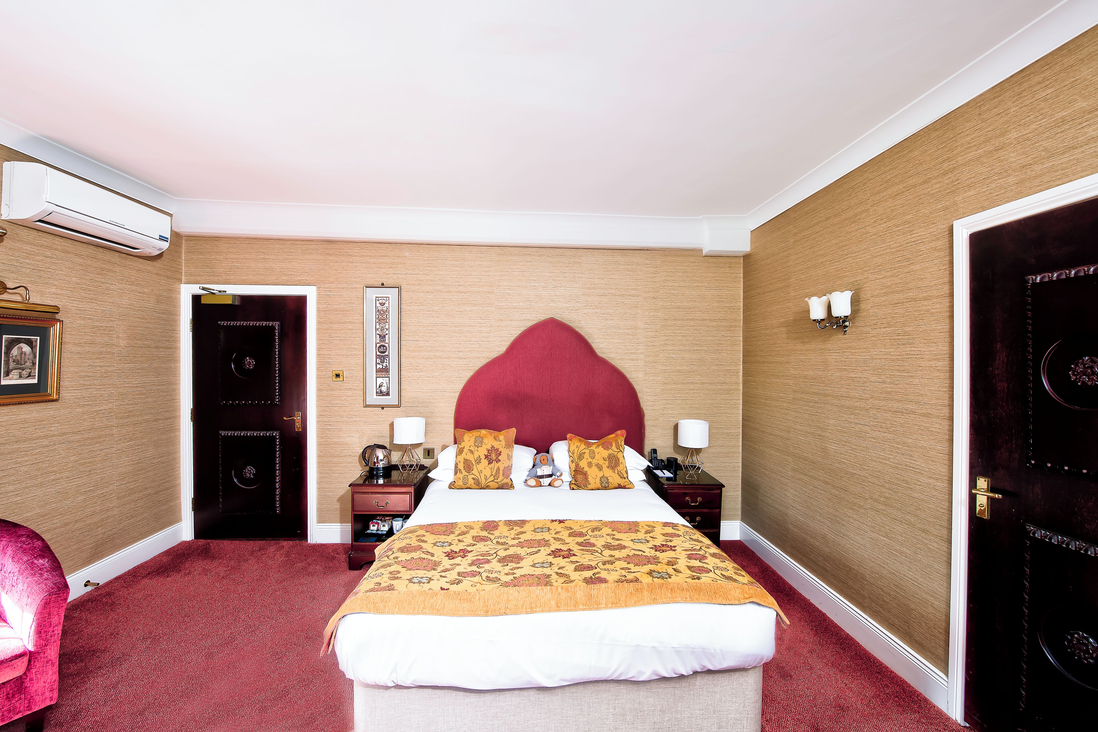 Quarto de hotel com uma cama grande, cabeceira vermelha, parede de madeira, uma porta e uma mesinha de cabeceira com uma luminária.