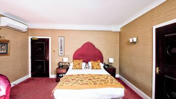 Quarto de hotel com uma cama grande, cabeceira vermelha, parede de madeira, uma porta e uma mesinha de cabeceira com uma luminária.