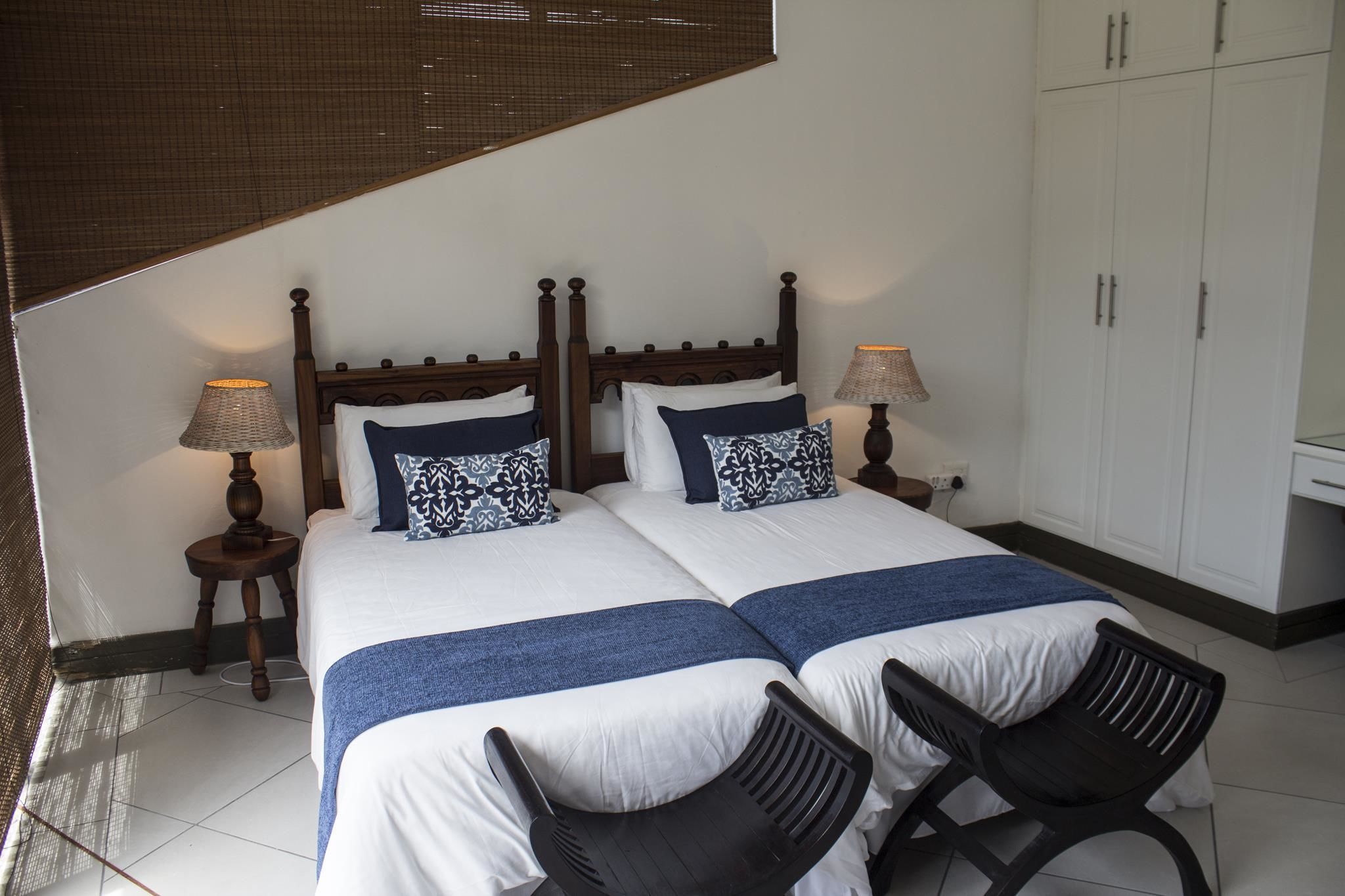 Uma cama de casal com cabeceira de madeira, duas mesas de cabeceira com luminárias, um cobertor azul e uma cadeira preta.