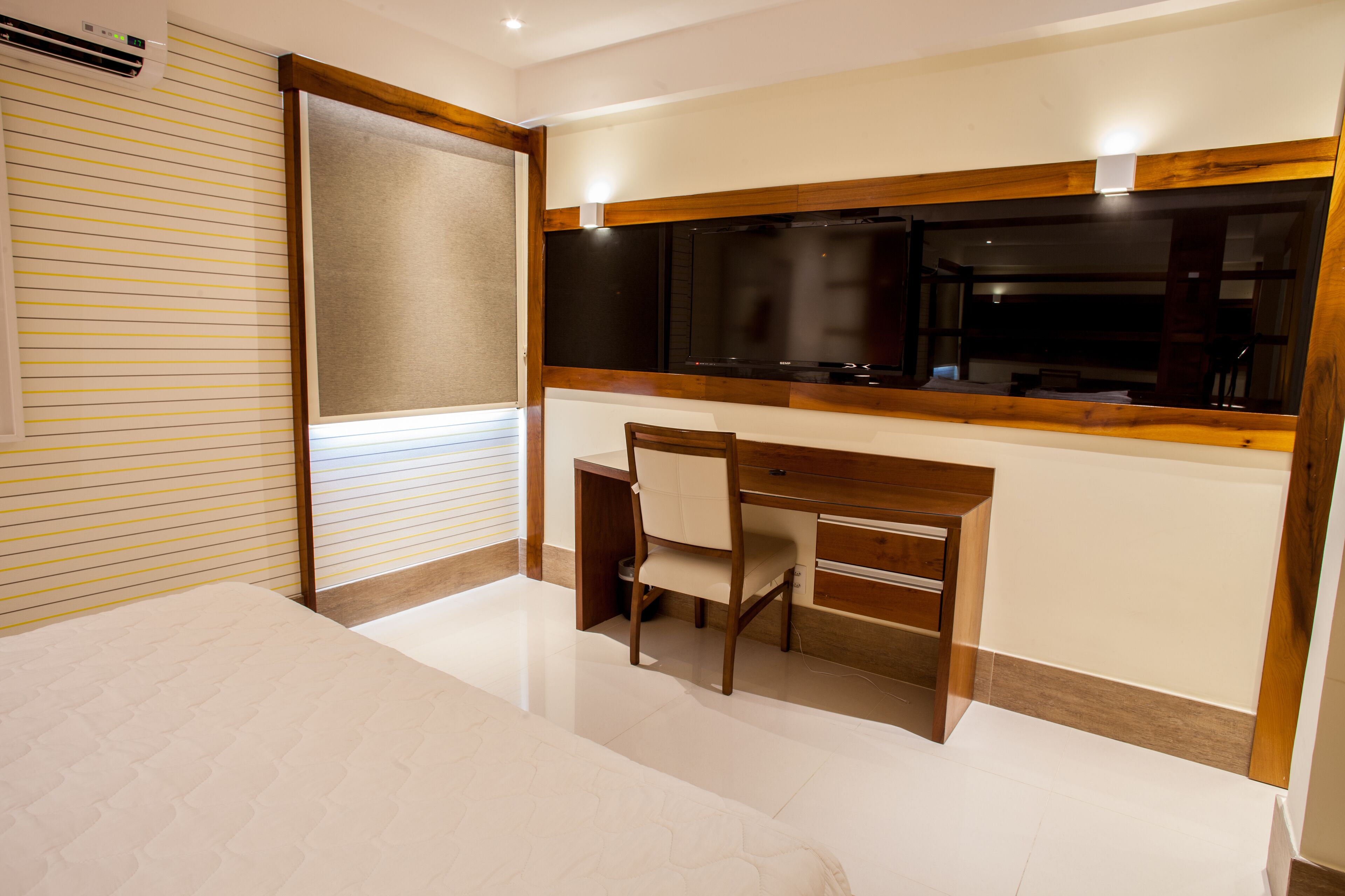 Quarto de hotel com cama, escrivaninha com cadeira, espelho grande e janela com persianas.