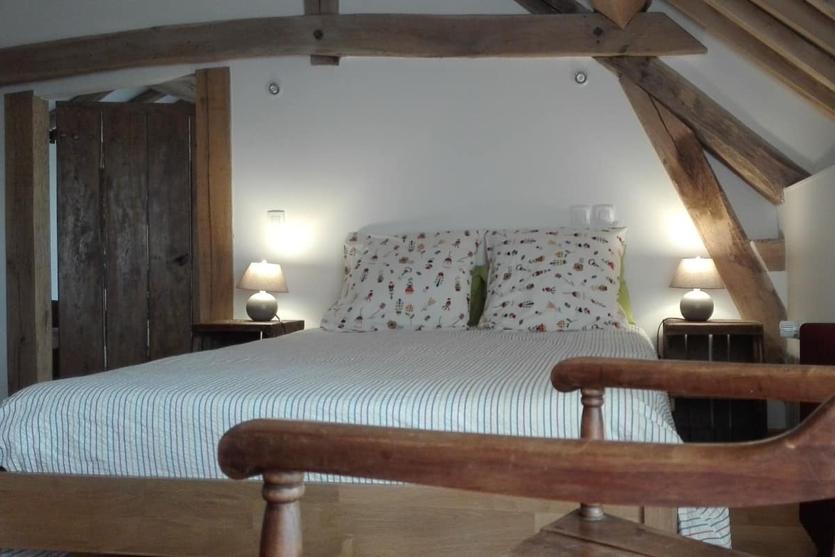Quarto com teto inclinado, cama com roupa de cama listrada, duas luminárias de cabeceira e uma cadeira de madeira.
