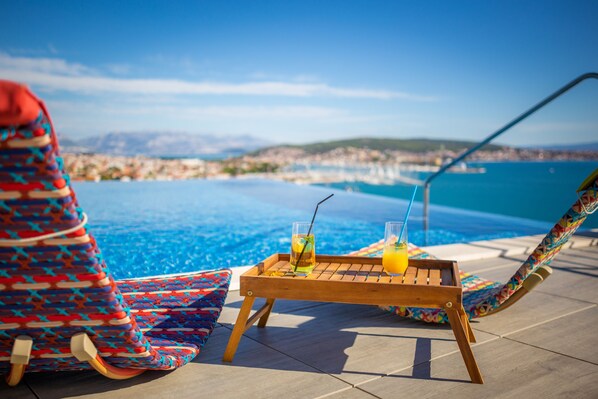 Área da piscina com mesa de madeira, duas bebidas e uma cadeira colorida.