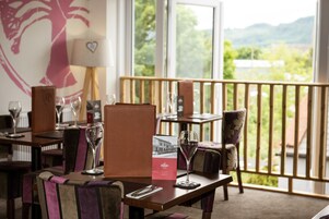 Restaurante com mesas preparadas para refeições, vista para o exterior e uma placa com o nome do restaurante.