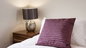 Cama bem arrumada com travesseiro branco e um roxo, mesa de cabeceira de madeira com luminária e gaveta.