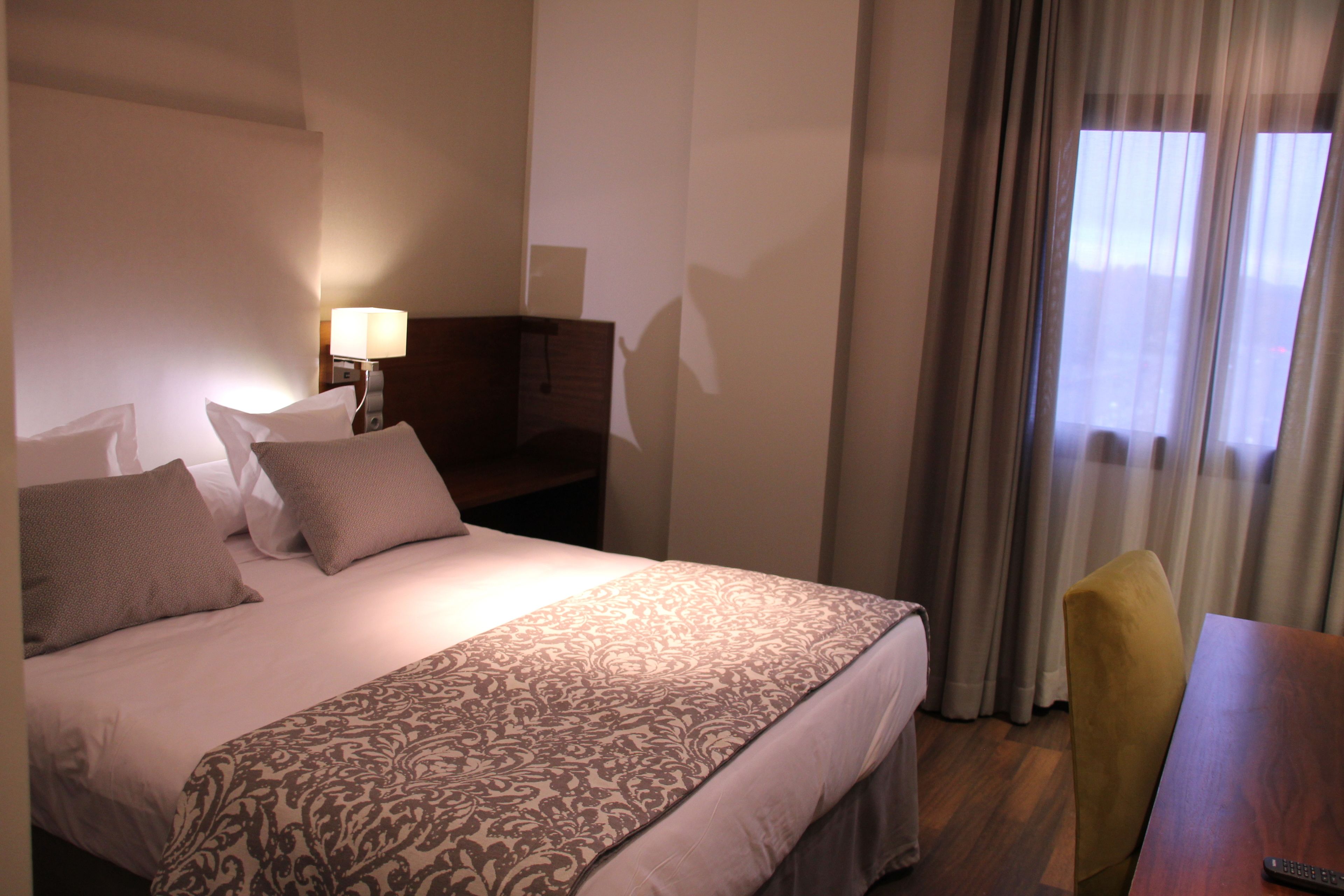 Quarto de hotel com cama, mesa de cabeceira, luminária, cabeceira de madeira, janela com cortinas e mesa de madeira.