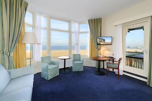 Quarto de hotel com uma grande janela dando vista para a praia, um sofá, duas poltronas, uma mesa redonda pequena e uma televisão.