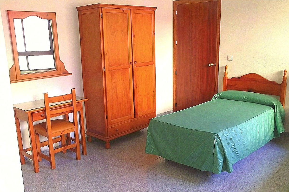 Quarto com escrivaninha de madeira, uma cadeira, guarda-roupa e cama com colcha verde.