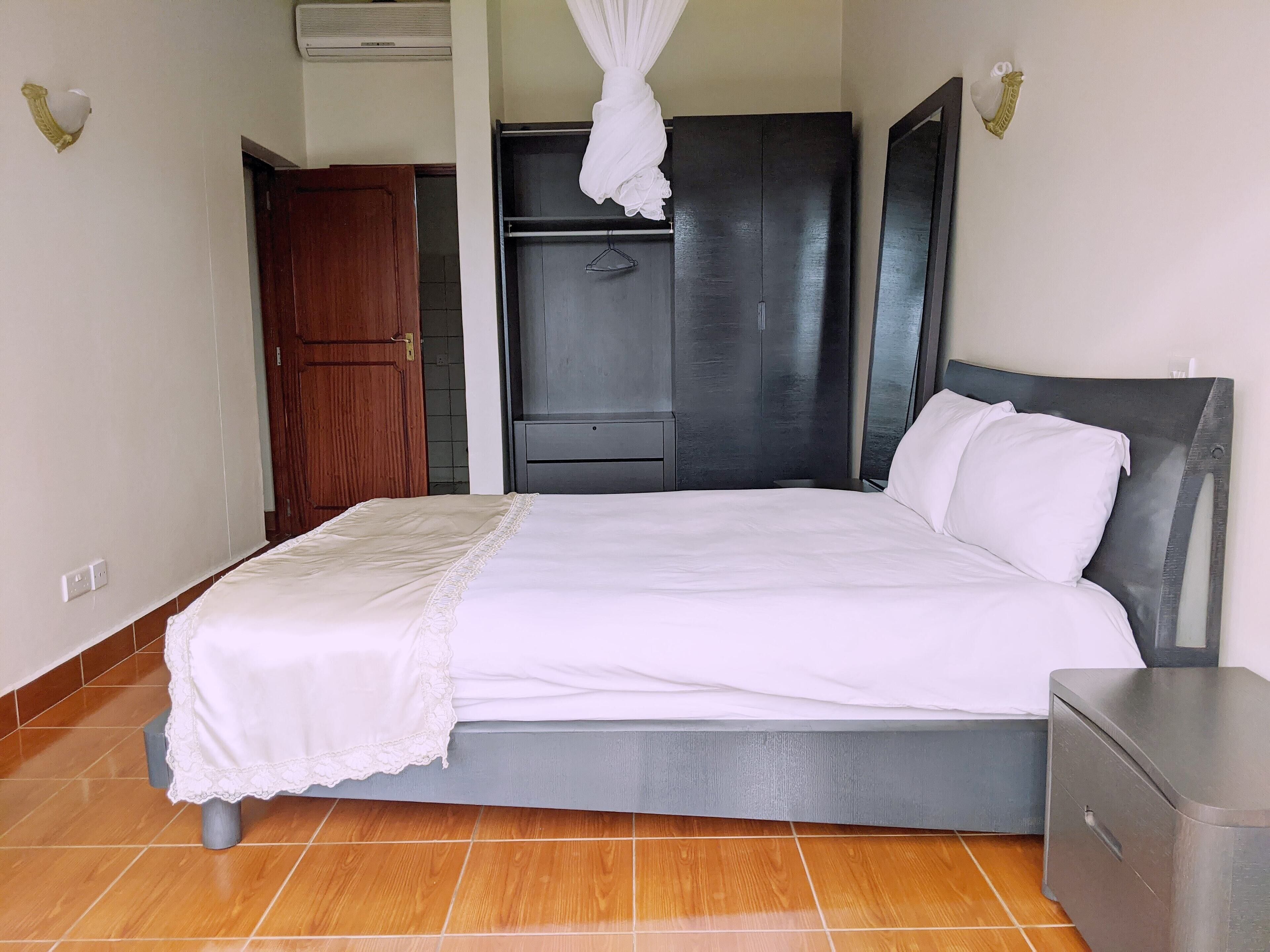 Uma cama bem arrumada com colcha branca e detalhes em renda, um guarda-roupa de madeira, uma pequena mesa de cabeceira e um ar-condicionado fixado na parede.