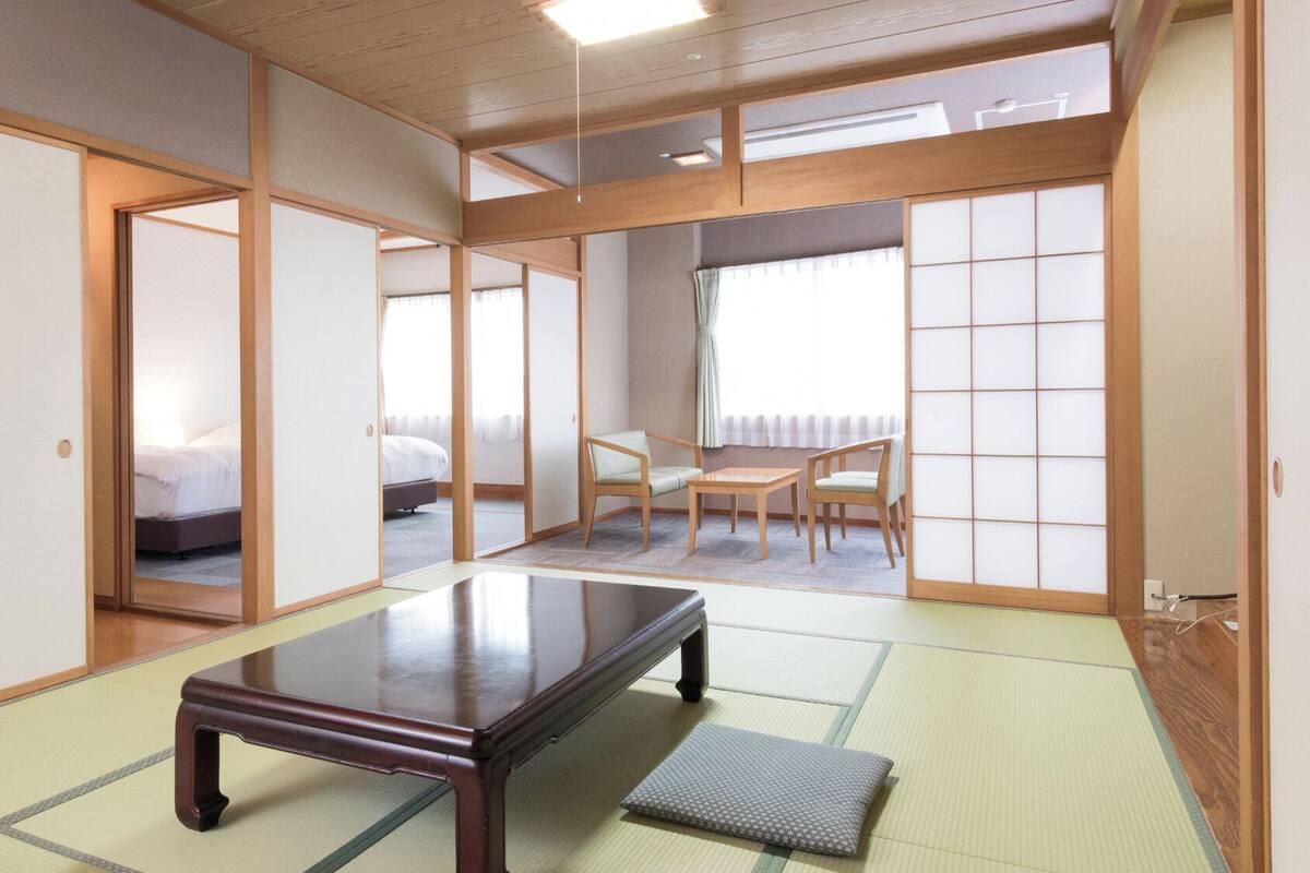 Um quarto japonês tradicional com tatames, uma mesa baixa, portas deslizantes e uma cama ao fundo.