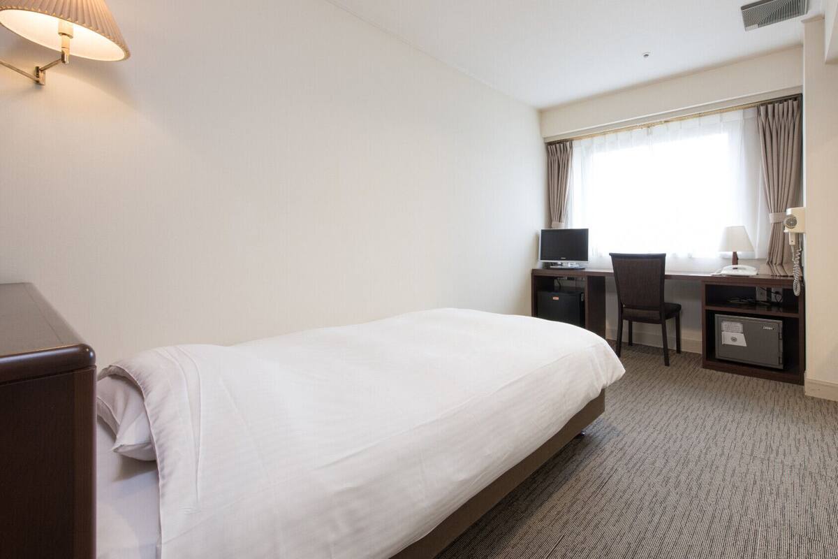 Quarto de hotel com cama, escrivaninha, cadeira e janela com cortinas.