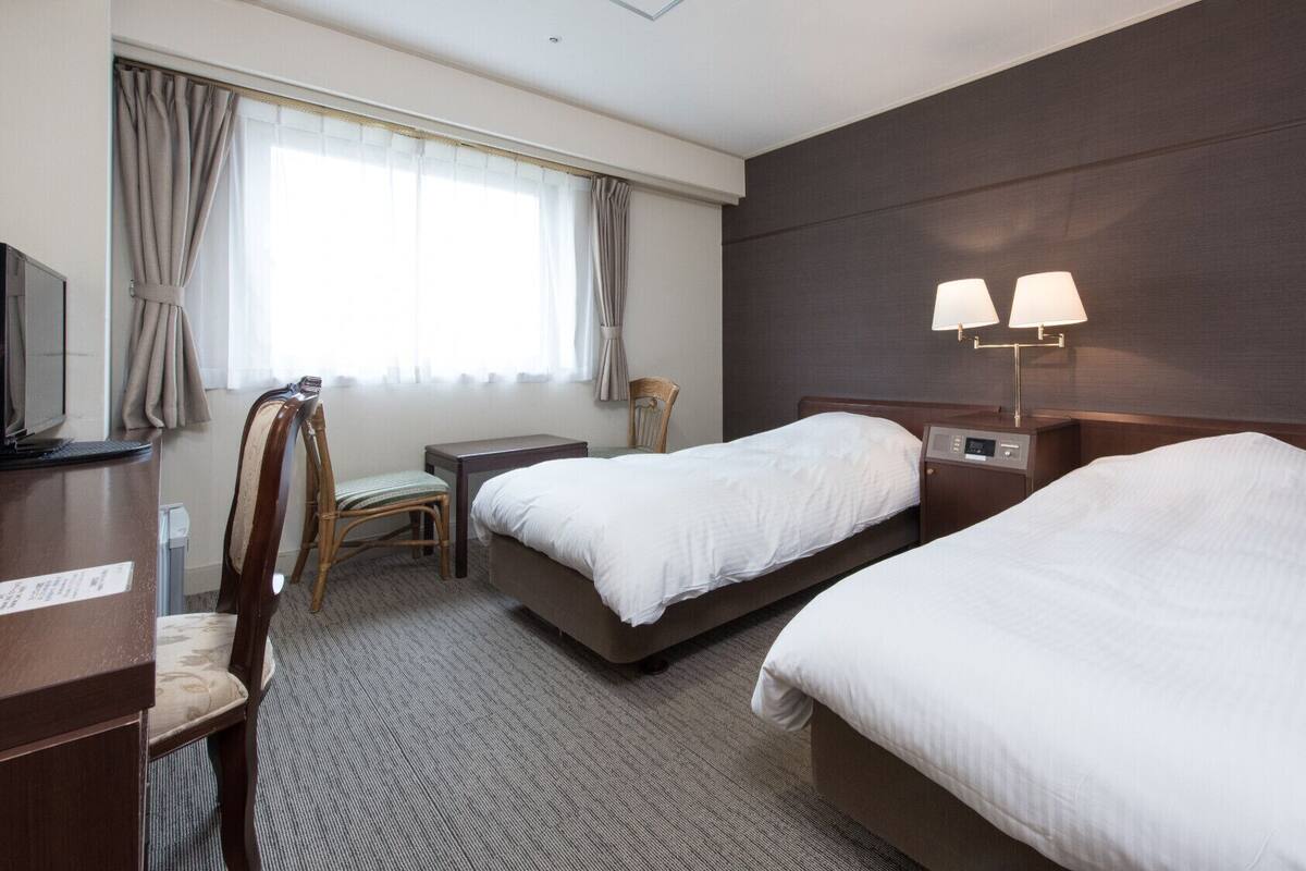 Quarto de hotel com duas camas, uma mesa de escritório com um computador, uma cadeira, uma luminária e uma janela com cortinas.