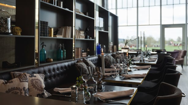 Um restaurante com uma longa mesa de jantar posta para uma refeição, um sofá de couro preto e prateleiras com livros e objetos decorativos.