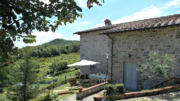 Um edifício de pedra com telhado ladrilhado, área de estar coberta ao ar livre e vista para uma paisagem verde exuberante.