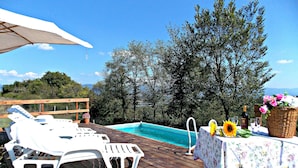 Um deck de madeira com piscina, cadeiras de lounge, uma mesa com toalha de mesa florida e uma cesta de vime com flores.