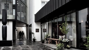 Um edifício moderno com grandes janelas, um pátio com plantas em vasos e uma escadaria que leva a um andar superior.