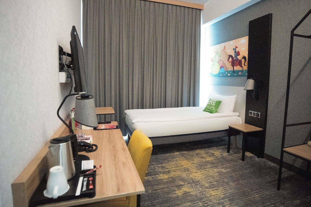 Quarto de hotel com cama, escrivaninha com computador e cafeteira, cadeira, quadro na parede e janela com cortinas.
