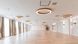 Corredor de hotel espaçoso, com piso de madeira, luminárias circulares no teto e grandes janelas com cortinas.