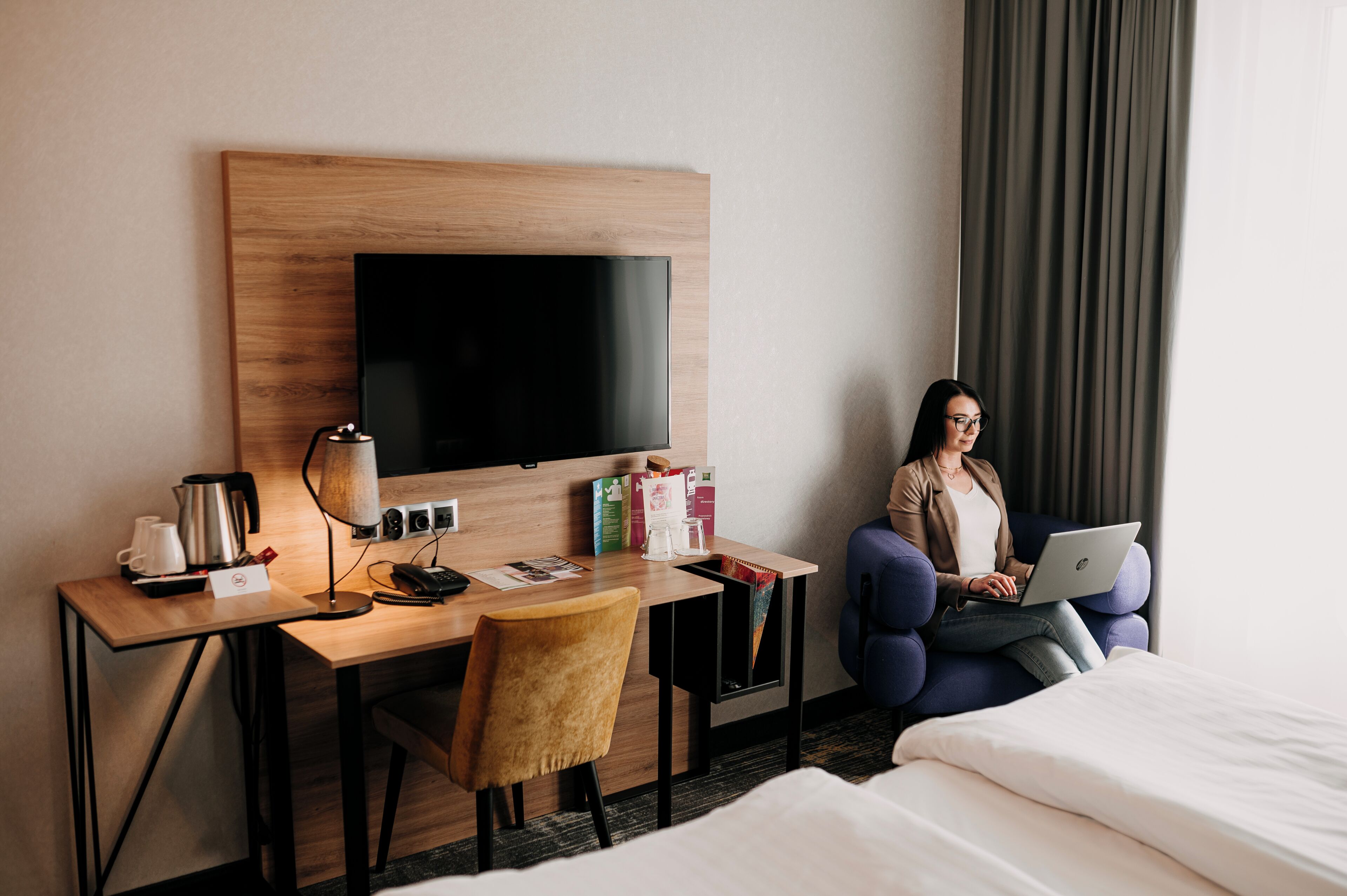 Quarto de hotel com escrivaninha de madeira, televisão de tela plana, laptop, cama e janela com cortinas.
