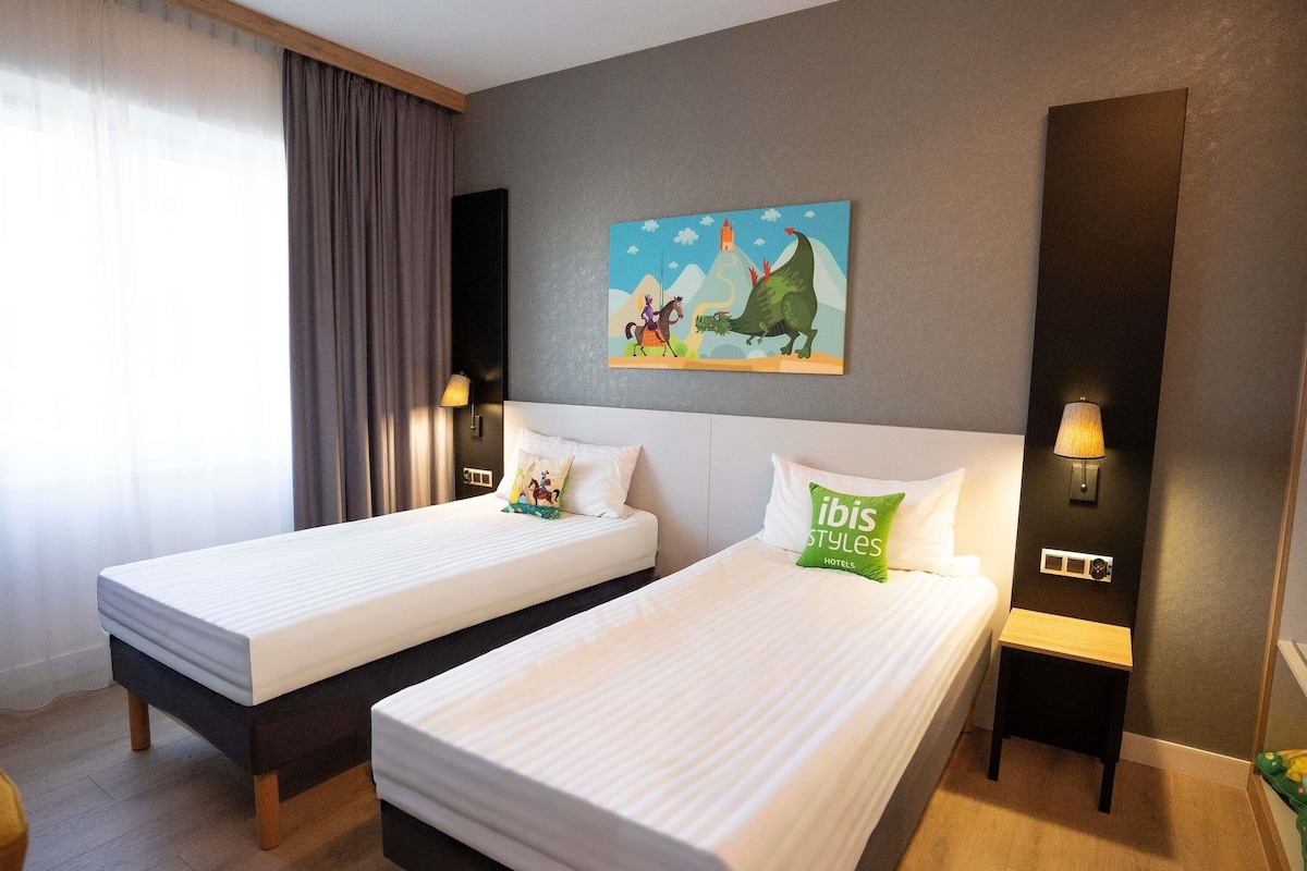 Quarto de hotel com duas camas, um quadro na parede, uma luminária e uma mesinha.