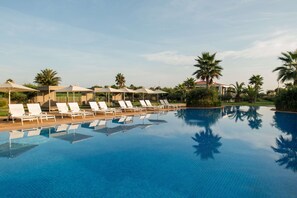 Piscina com espreguiçadeiras brancas e guarda-sóis, cercada por palmeiras e um céu claro.