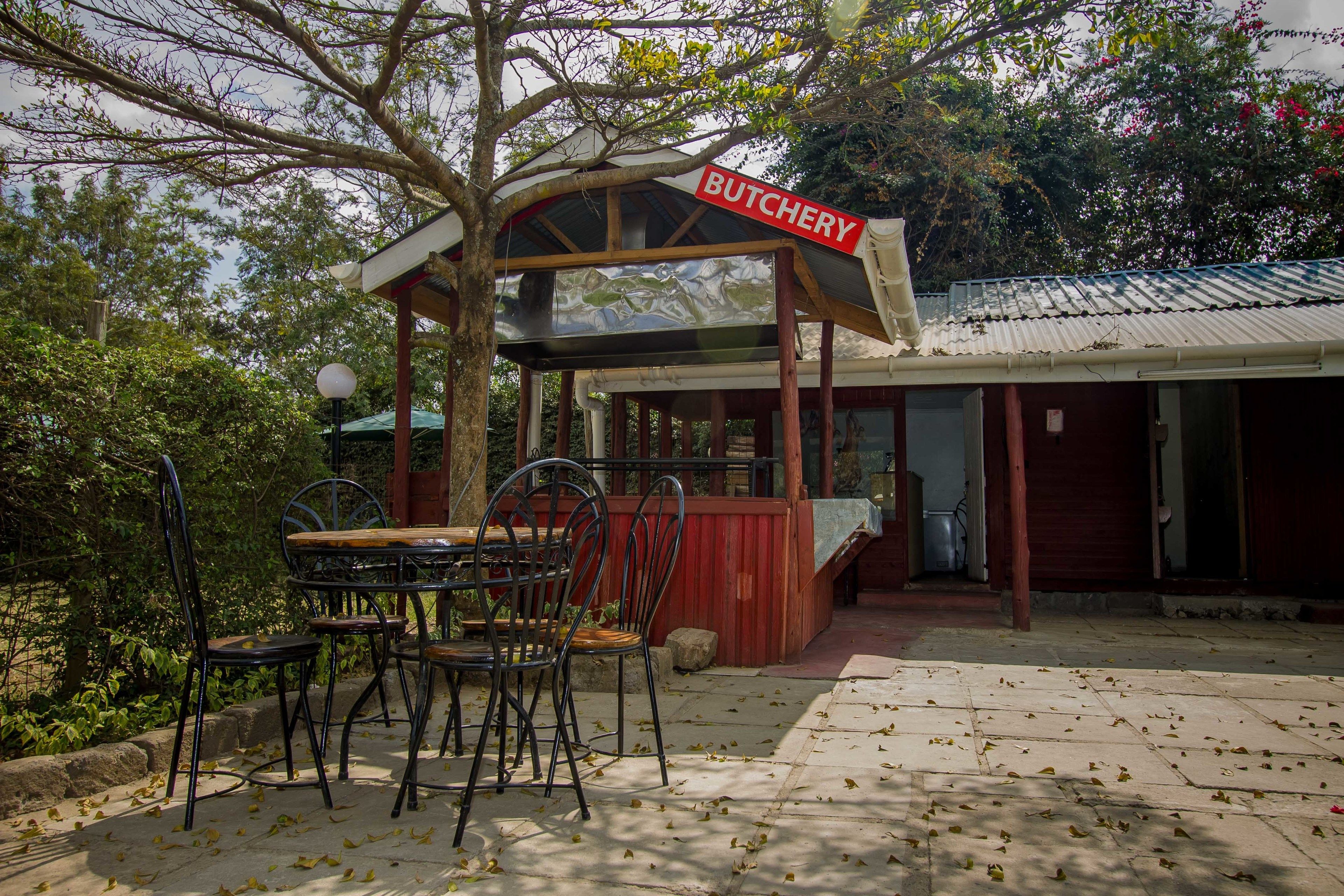 Uma área de estar rústica ao ar livre, com uma mesa e cadeiras, uma placa com a inscrição 'Butchery' e um edifício com telhado de metal corrugado.