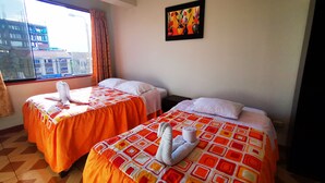 Quarto de hotel com duas camas, cada uma com colcha laranja e almofadas brancas, e uma janela com vista para prédios.