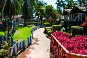 Uma passagem serpenteia por um jardim exuberante, com uma cerca de madeira de um lado e um edifício com telhado de palha do outro.