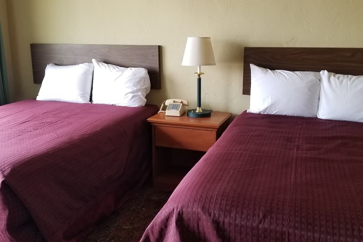 Quarto de hotel com duas camas, cada uma com cabeceira de madeira, almofadas brancas e cobre roxa. Há uma mesa de cabeceira com abajur e telefone.