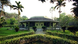 Um edifício com telhado verde e um caminho pavimentado que leva a ele, rodeado por vegetação bem cuidada e palmeiras.