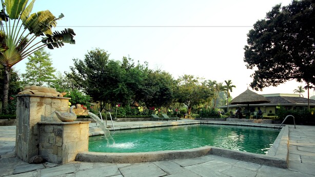 Uma piscina com uma fonte de pedra, rodeada por azulejos de pedra e vegetação exuberante.