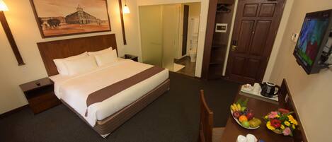 Quarto de hotel com cama, escrivaninha com fruteira, máquina de café e televisão.