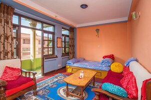 Quarto com sofá, mesa de madeira, cama com roupa de cama colorida, tapete estampado e janelas amplas com cortinas.