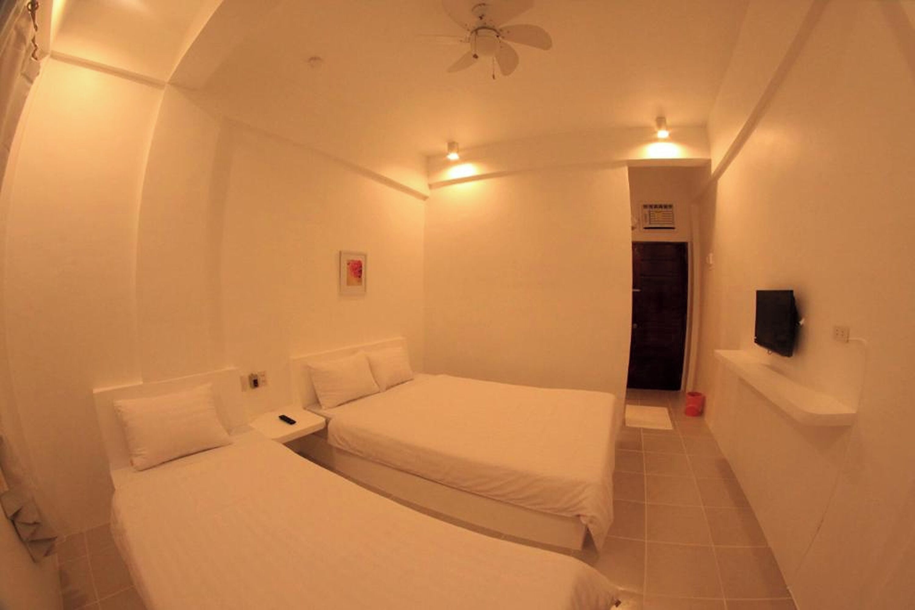 Quarto com duas camas, um ventilador de teto, uma televisão pequena numa prateleira e uma porta para um banheiro.