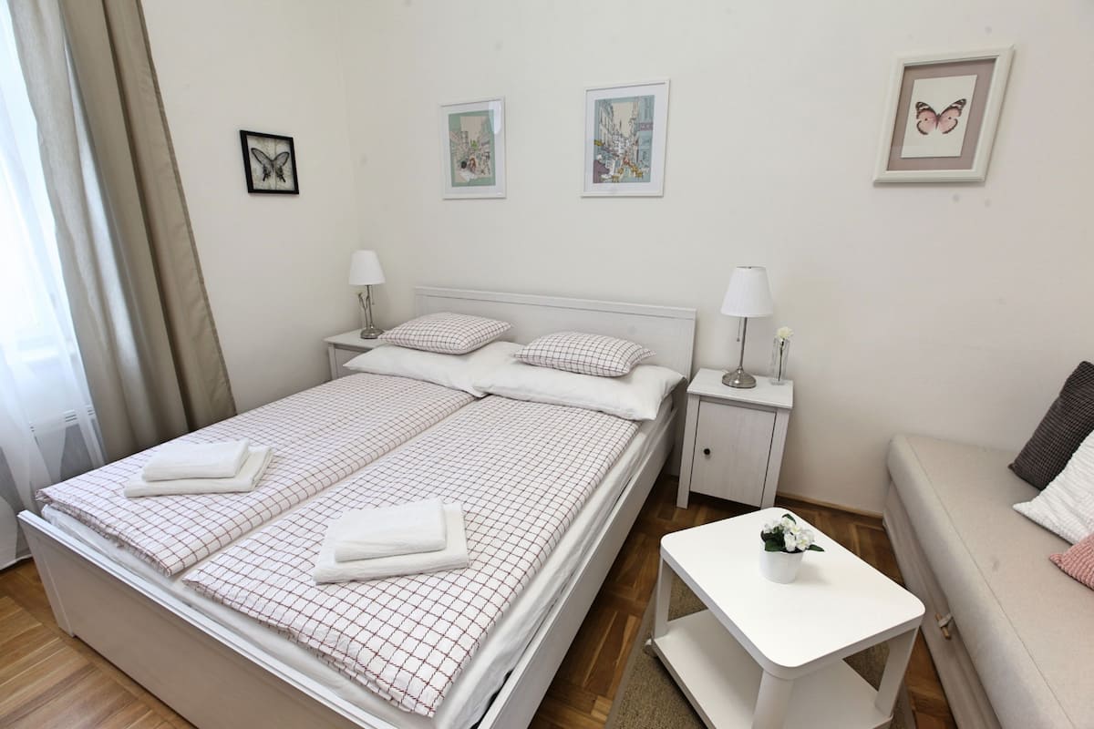Quarto bem arrumado com uma cama de casal, duas mesas de cabeceira com luminárias, uma mesinha branca com um vaso e um sofá com almofadas.