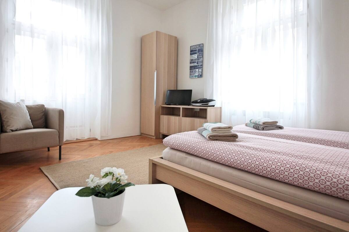 Quarto de hotel moderno com cama, sofá, escrivaninha com um laptop e um vaso de planta.