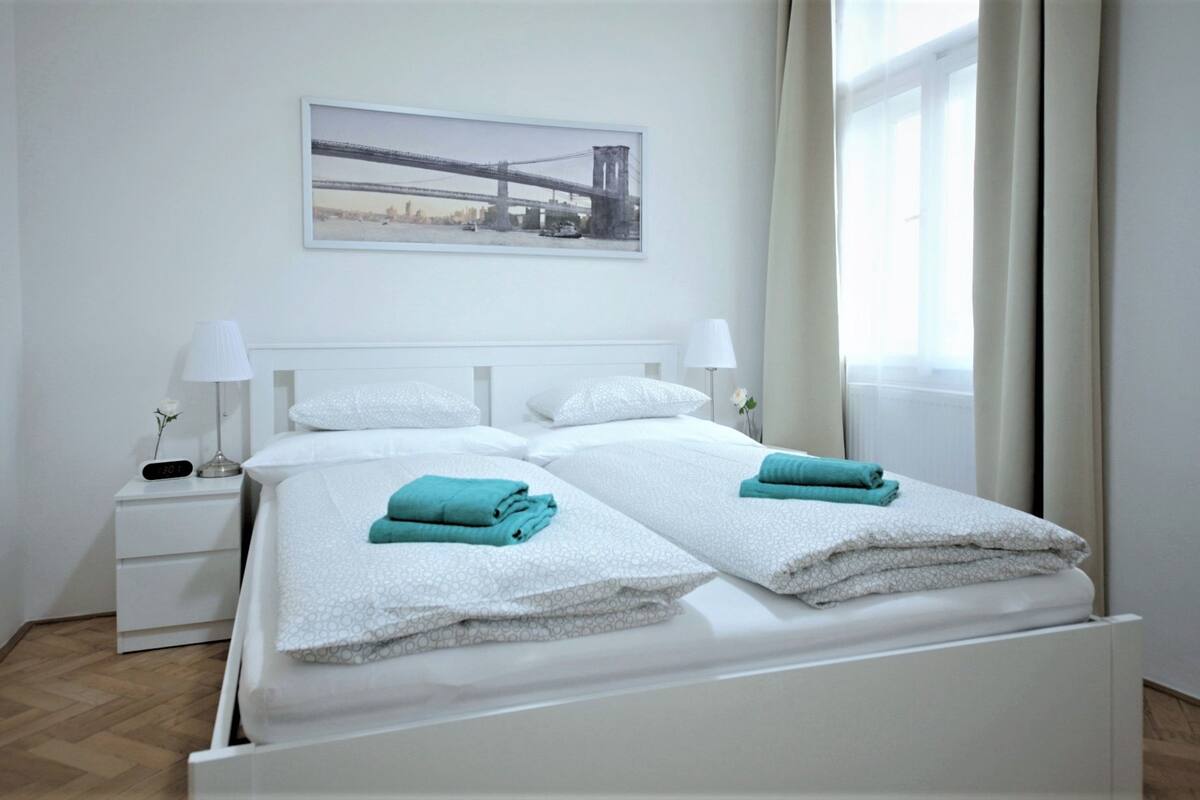 Uma cama de casal com roupa de cama branca e toalhas verde-água, uma mesa de cabeceira com abajur, um quadro na parede e uma janela com cortinas.
