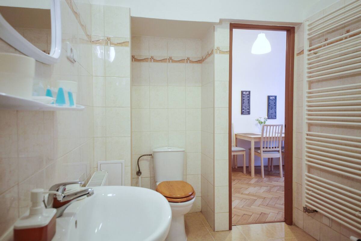Casa de banho com sanita, lavatório e espelho. Uma porta dá para uma área de refeições com mesa e cadeiras.