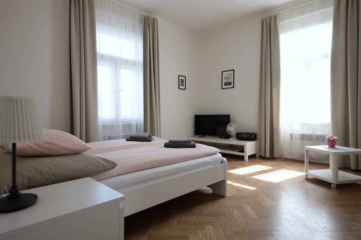 Quarto de hotel moderno com uma cama, uma TV, uma mesinha de cabeceira e uma pequena mesa com uma flor.