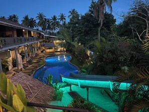 Uma área de piscina em resort, com espreguiçadeiras e guarda-sóis, rodeada por vegetação exuberante e palmeiras.