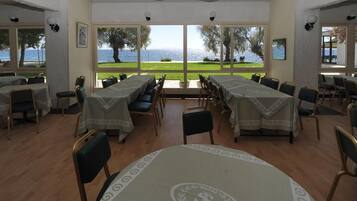 Uma área de jantar com mesas e cadeiras, janelas amplas com vista para o mar e árvores, e piso de madeira.