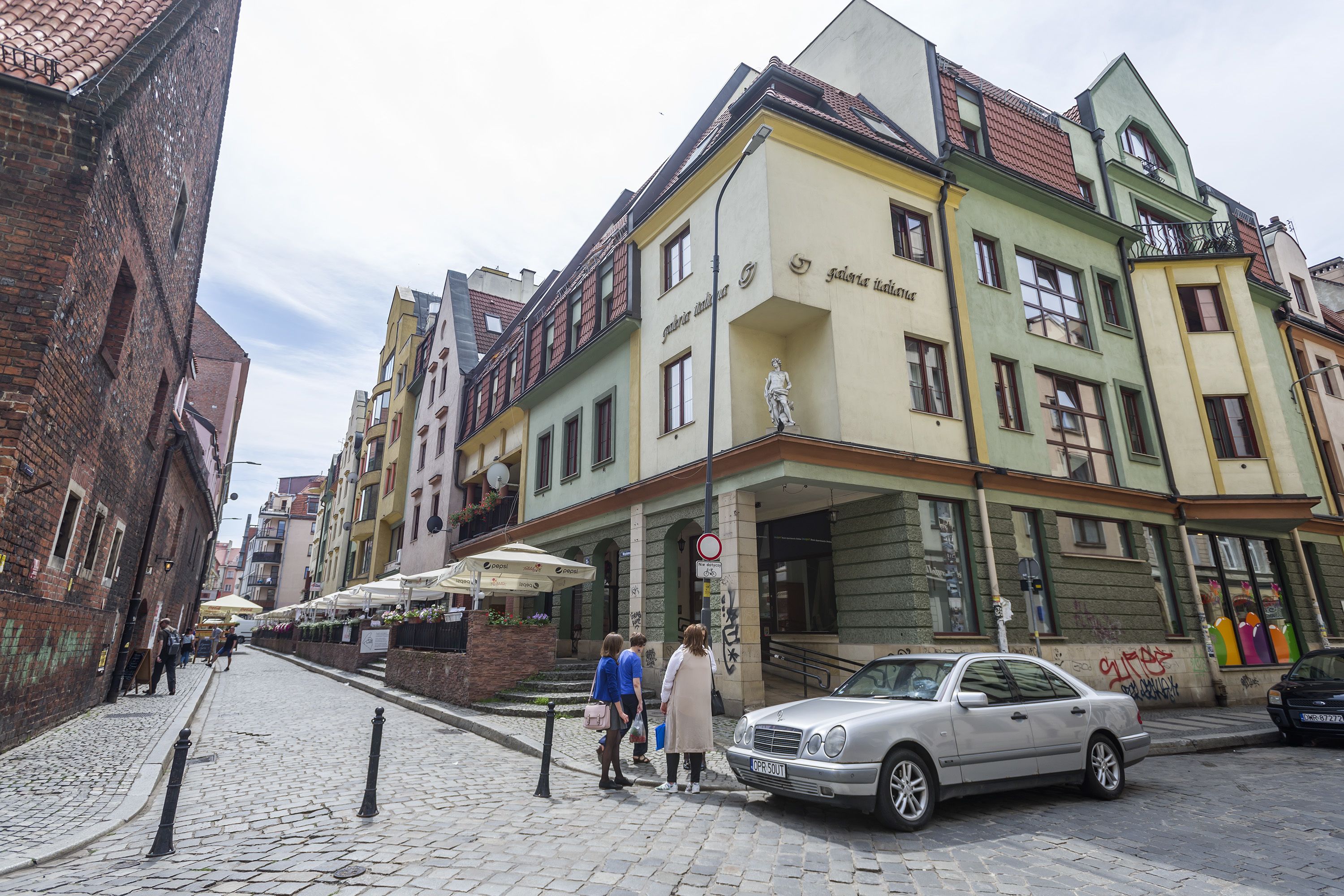 Rua de paralelepípedos com um edifício que apresenta uma estátua, mesas ao ar livre e um carro estacionado.