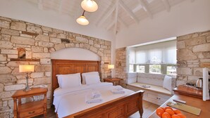 Quarto com parede de pedra, cama de madeira, abajures, um assento junto à janela e uma tigela de laranjas sobre a mesa.