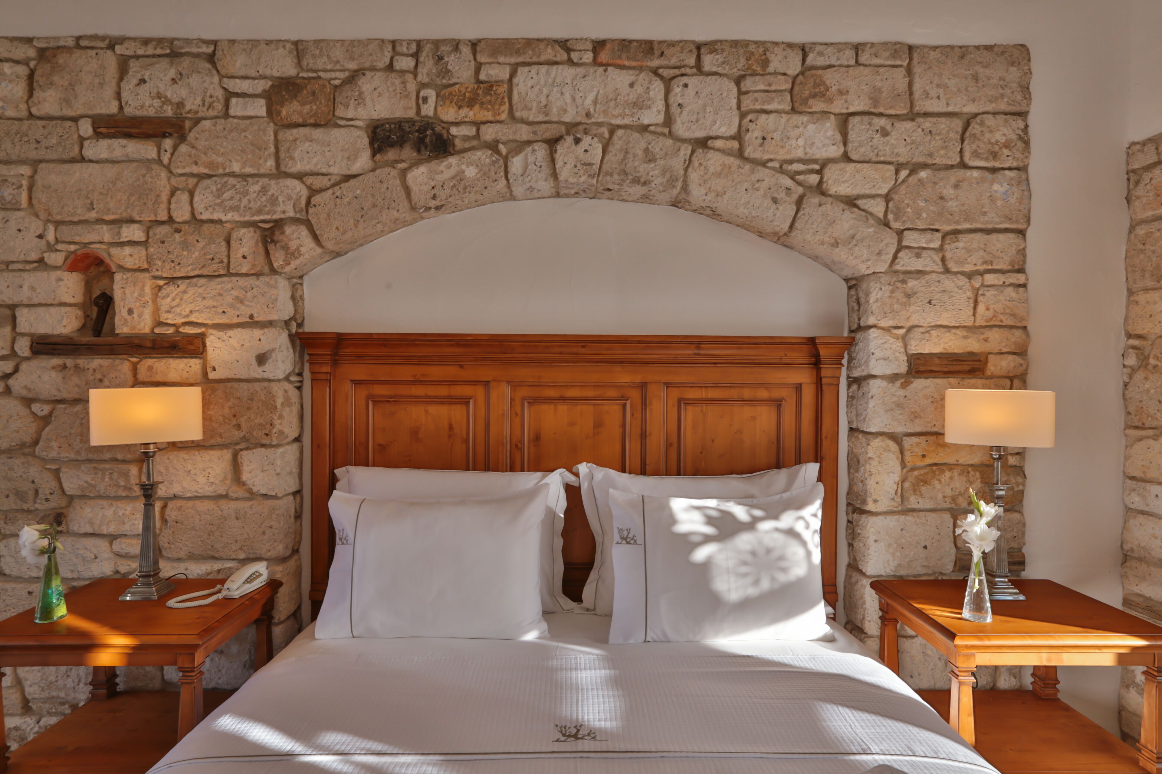 Quarto de hotel com parede de pedra, cabeceira de madeira, duas mesas de cabeceira com luminárias e uma cama com lençóis brancos.