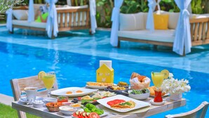 Disposição para café da manhã à beira da piscina, com diversos pratos, bebidas e um recipiente em formato de casa amarela.
