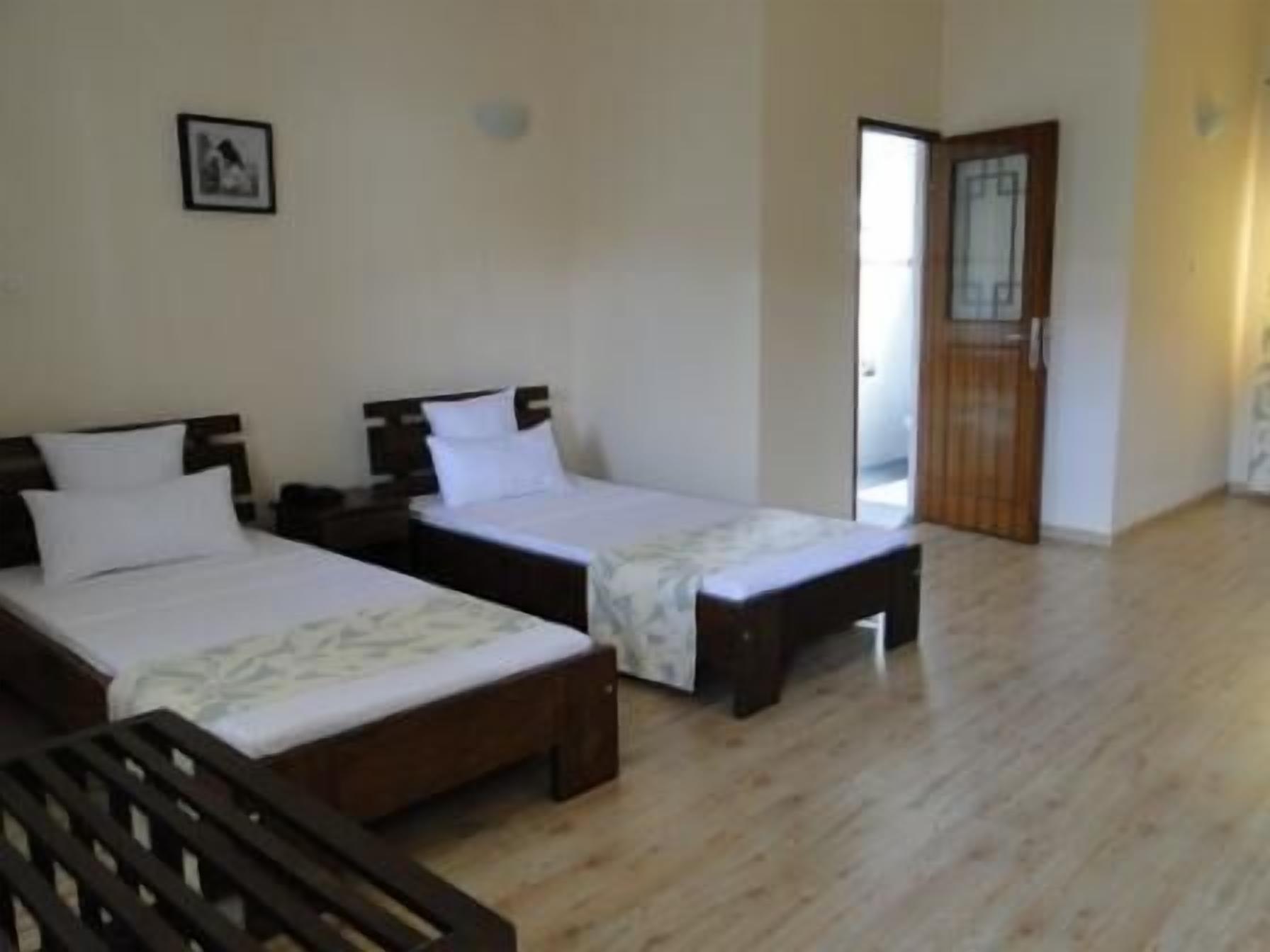 Quarto com duas camas de solteiro, um banco de madeira, uma mesa pequena e uma porta para o exterior.