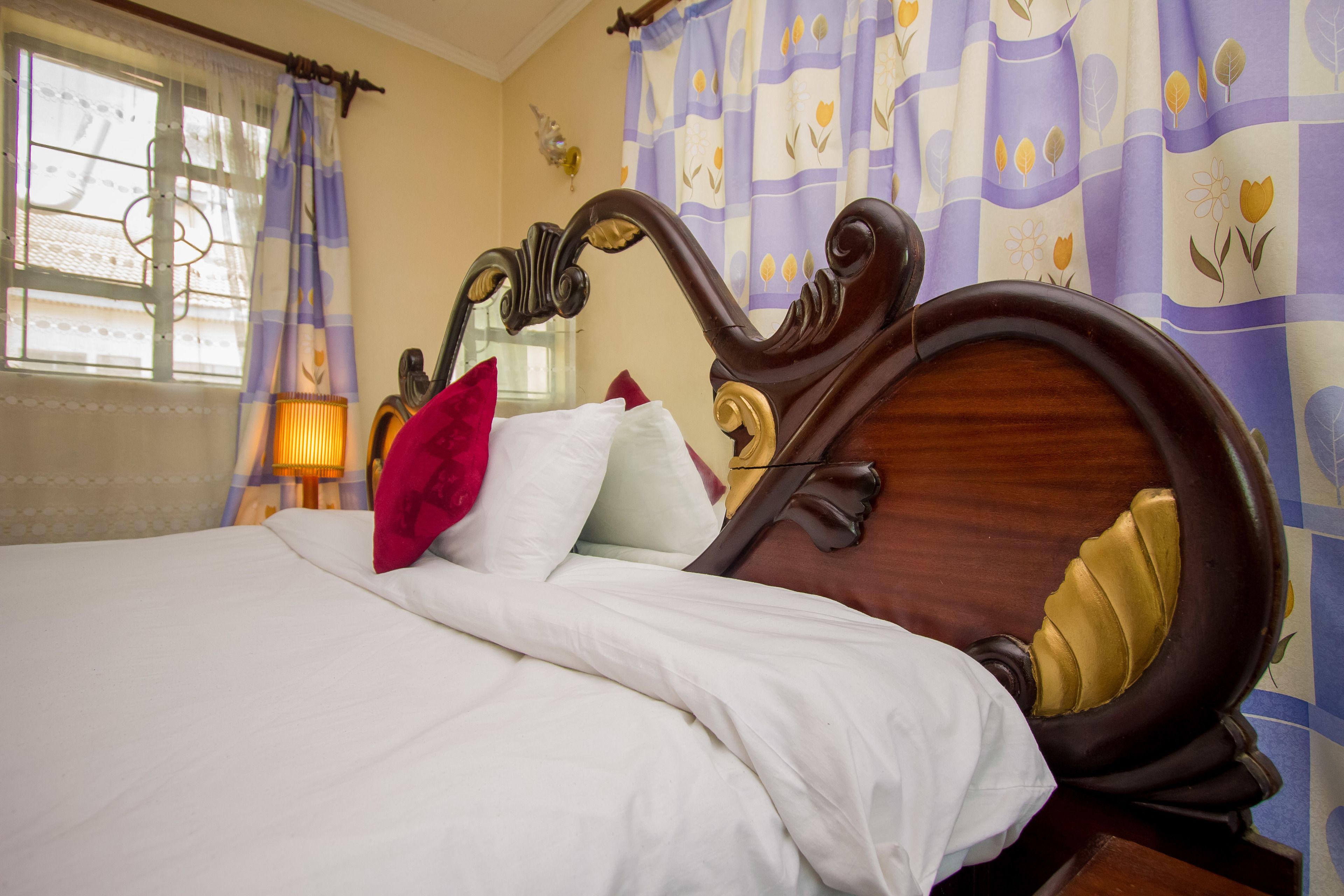 Uma cama bem arrumada com lençóis brancos e um travesseiro vermelho, uma cabeceira de madeira e uma janela com cortinas.
