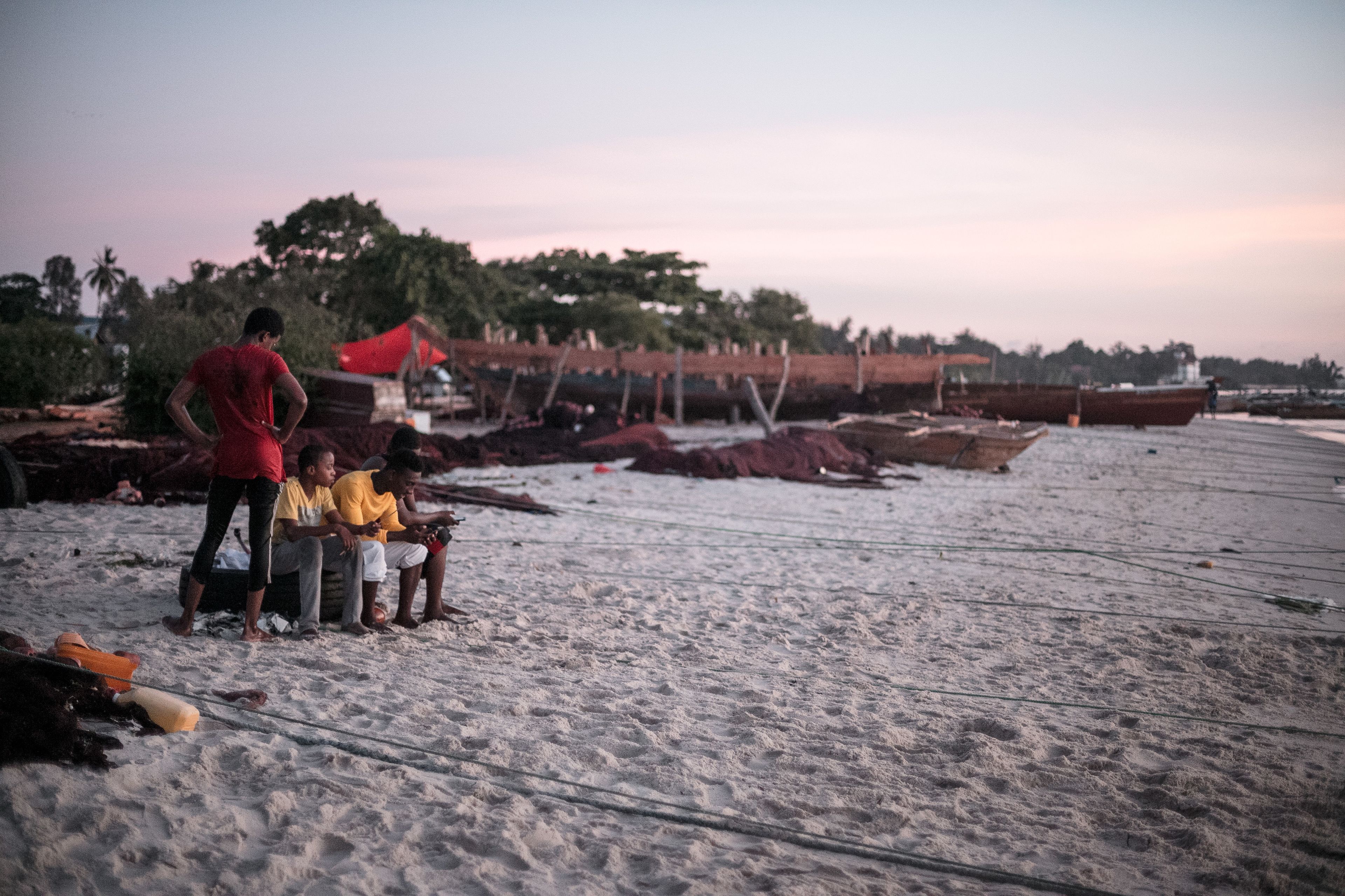 Uma praia arenosa com pessoas sentadas e em pé, alguns barcos ao fundo e uma estrutura vermelha.