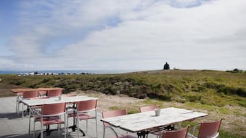Uma área externa com mesas e cadeiras, com vista para uma praia e dunas.